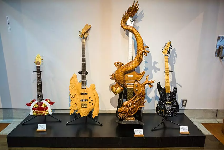 Štyri elektrické gitary sa opierajú o stojany, každá so zložitým dreveným dizajnom. Jedným z nich je prepracovaný drak, ktorý siaha až po krk gitary